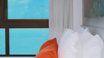 Quarto com uma janela ampla, roupa de cama branca e vista para o oceano.