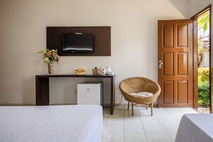 Quarto de hotel com cama, escrivaninha com televisão, cadeira de vime e porta que dá para uma área externa.