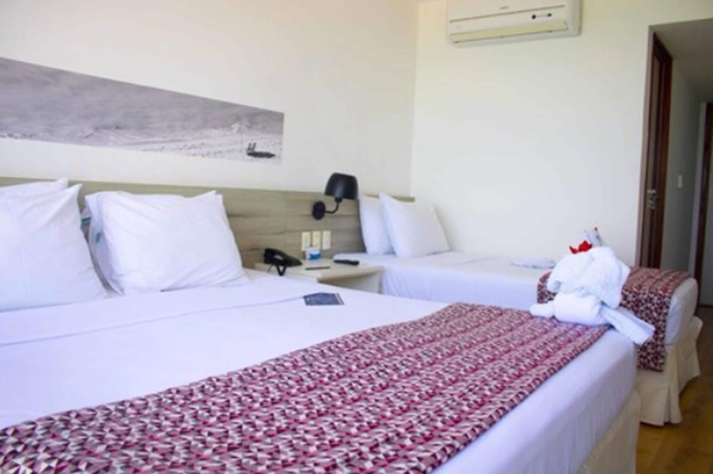 Quarto de hotel com duas camas, uma mesa de cabeceira com abajur, telefone e um quadro na parede.