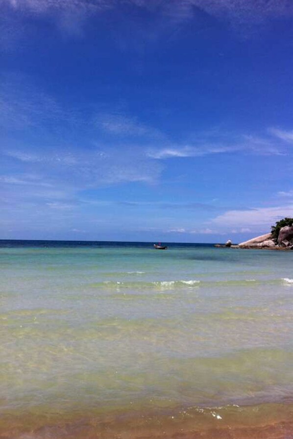 Céu azul límpido sobre um mar tranquilo, com um barco ao longe e terreno rochoso à direita.
