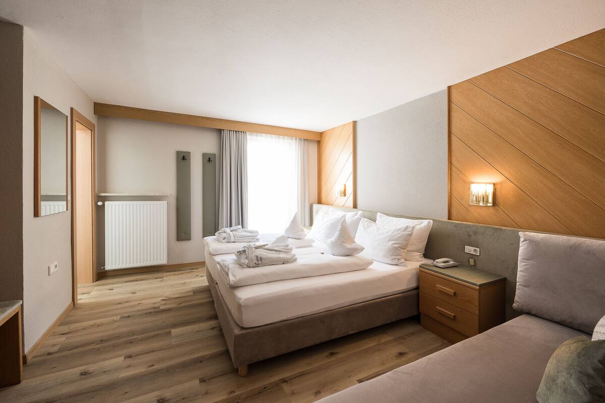 Quarto de hotel com cama, mesa de cabeceira e criado-mudo. Há uma janela com cortinas, um radiador e um painel de parede em madeira.
