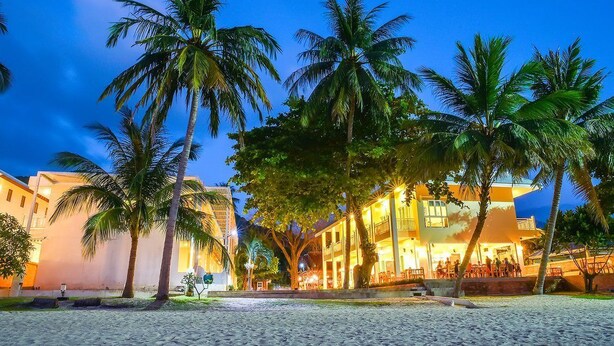 Um hotel à beira-mar com palmeiras, uma praia de areia e um edifício com várias varandas e área para sentar ao ar livre.