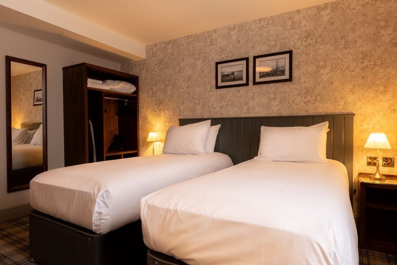 Quarto de hotel com duas camas, um guarda-roupa, um espelho e duas imagens emolduradas na parede.