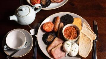 Mesa de café da manhã com pratos de ovos, salsichas, tomates, cogumelos, feijão, torradas e presunto.