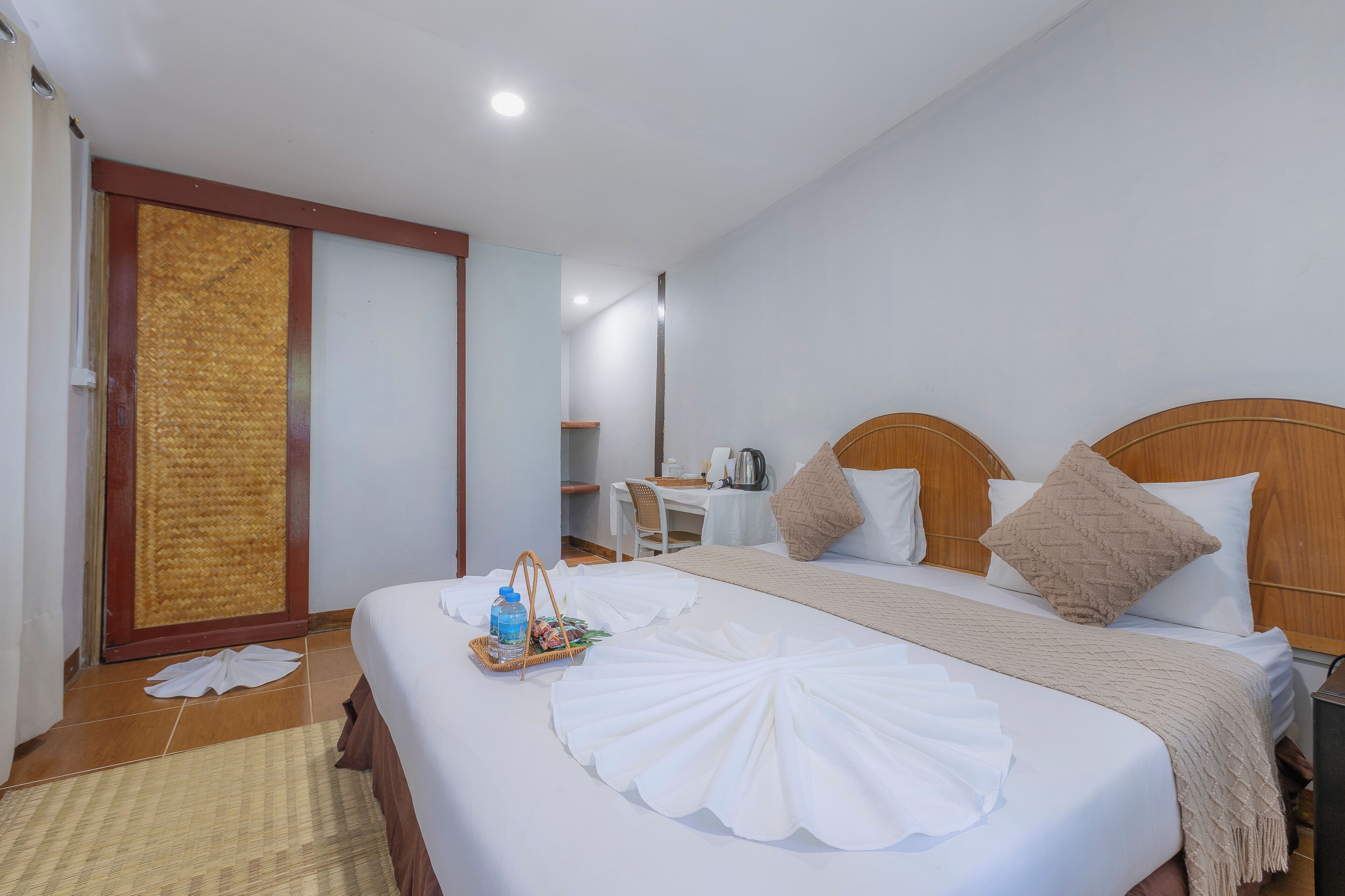 Quarto de hotel com cama bem arrumada, cabeceira de madeira, uma mesa pequena com chaleira e escrivaninha com cadeira.