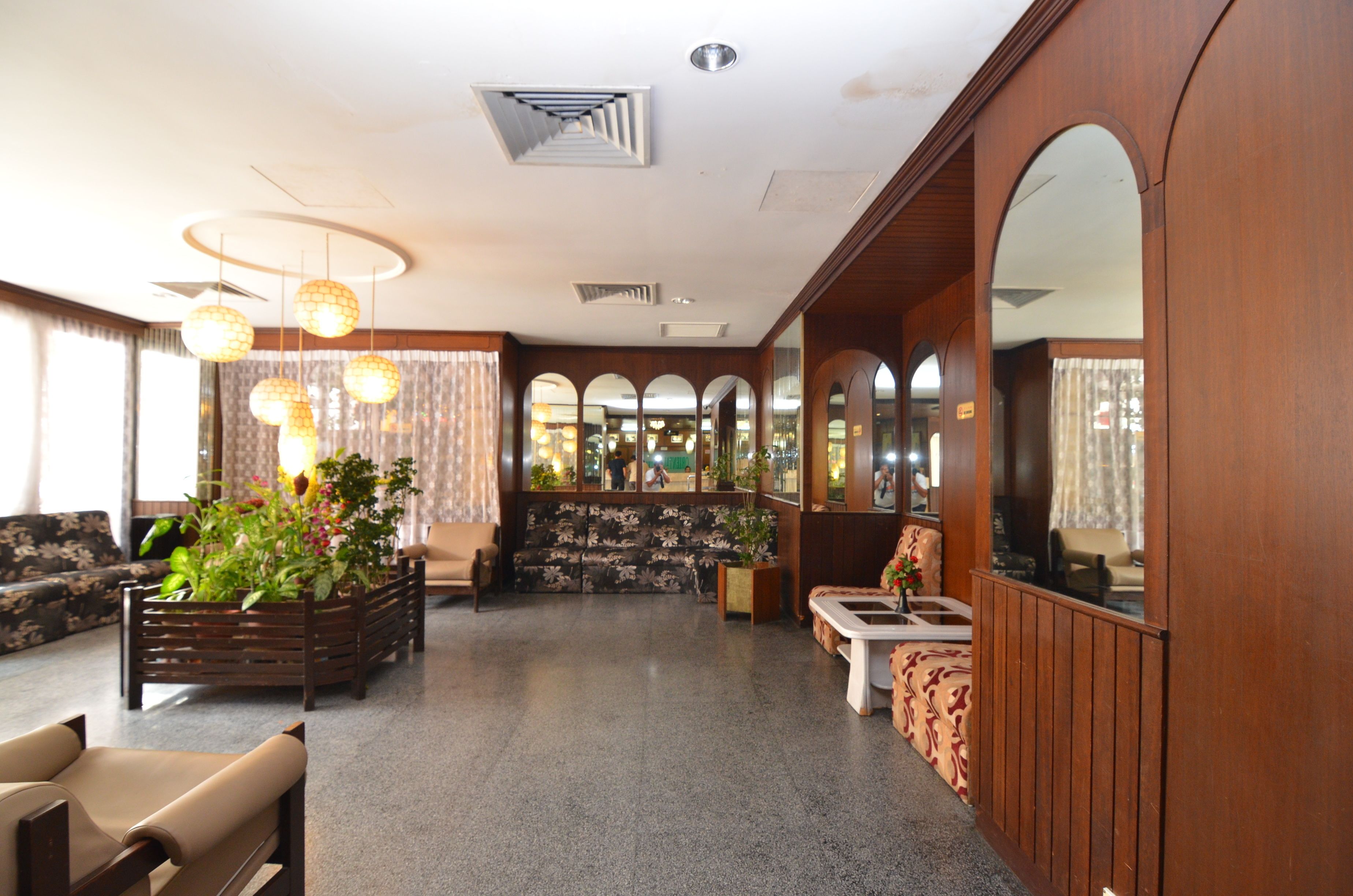 Um hall amplo com um balcão de recepção em madeira, área de estar com sofás e um espelho grande que reflete o interior.