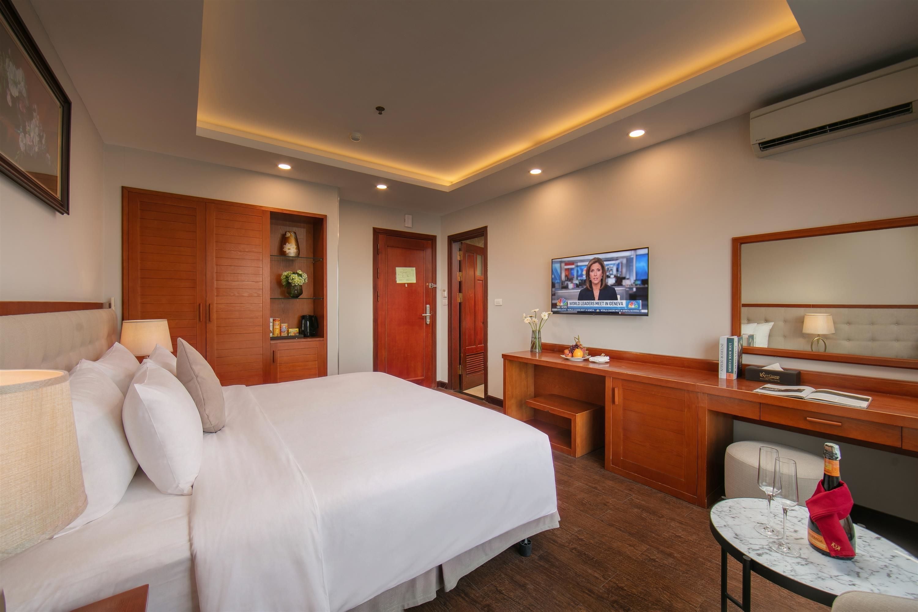 Quarto de hotel com cama, escrivaninha, televisão e pequena mesa com garrafa e copos.