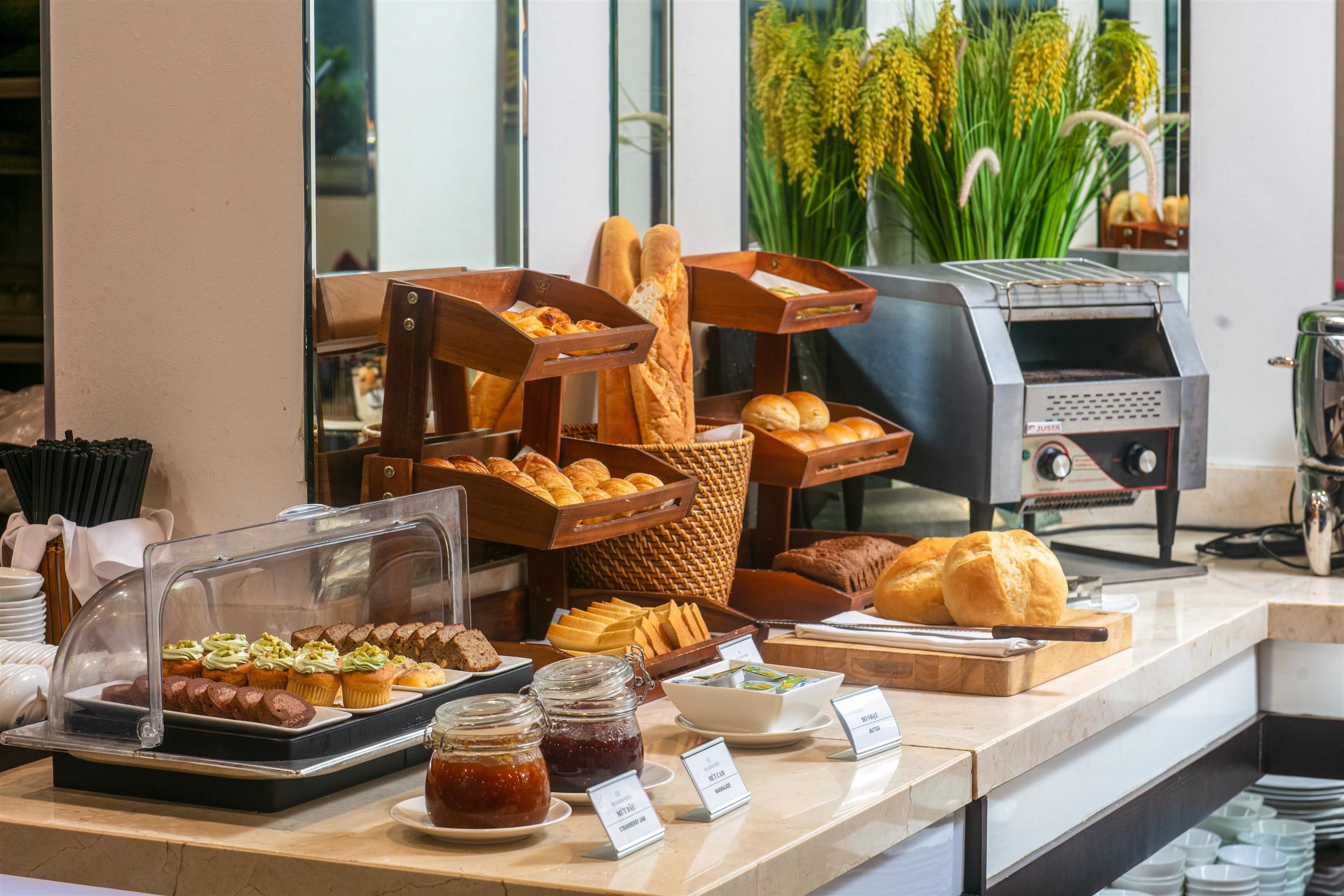 Buffet variado com produtos de panificação, incluindo pão, muffins e pastelaria, com máquina de café e expositor de acompanhamentos.