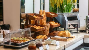 Buffet variado com produtos de panificação, incluindo pão, muffins e pastelaria, com máquina de café e expositor de acompanhamentos.