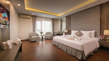 Quarto de hotel com cama grande, duas poltronas, uma mesa pequena com flores e um espelho amplo.