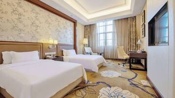 Quarto de hotel com duas camas, uma escrivaninha, uma cadeira, uma televisão e uma janela grande com cortinas.