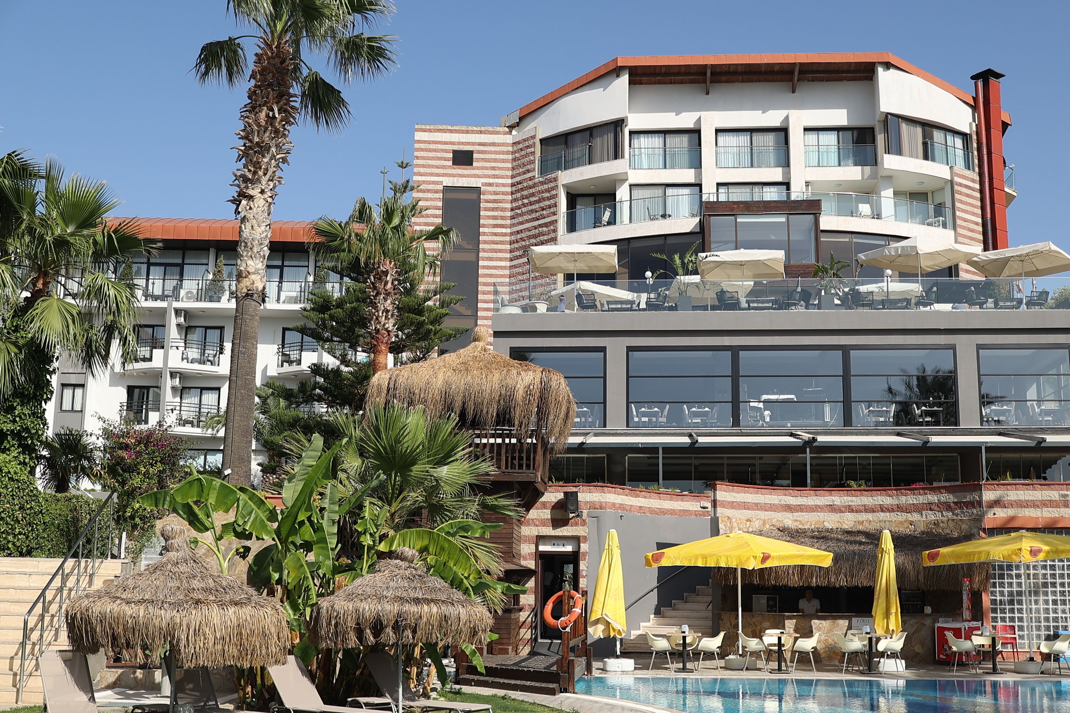 Um hotel com área de piscina, guarda-sóis de palha e um edifício de vários andares com varandas.