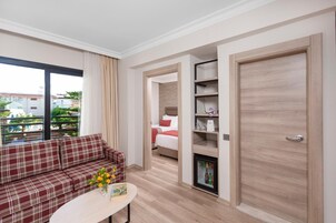 Quarto de hotel moderno com sofá xadrez, uma pequena mesa com um vaso de flores, uma cama com roupa de cama vermelha e estantes embutidas com um minibar.