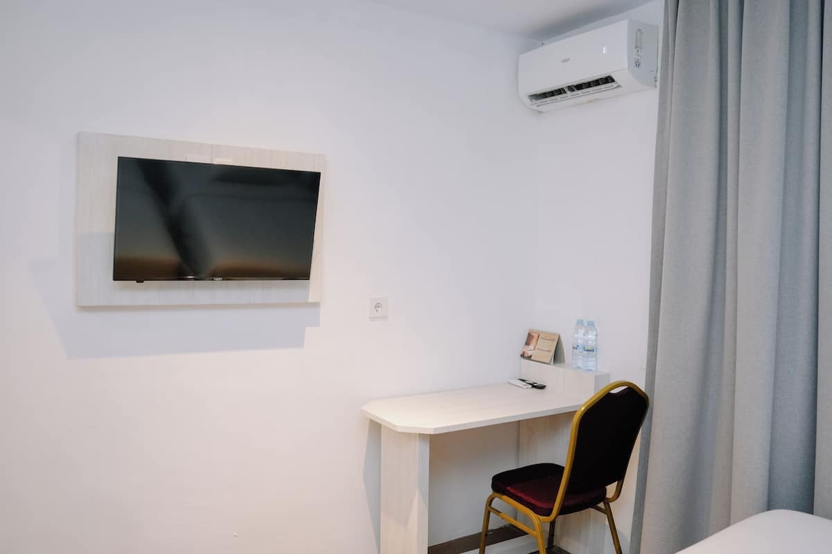 Quarto de hotel com escrivaninha branca, cadeira, televisão de tela plana, ar condicionado, mesinha com porta-retrato e garrafas de água, e cortina cinza.