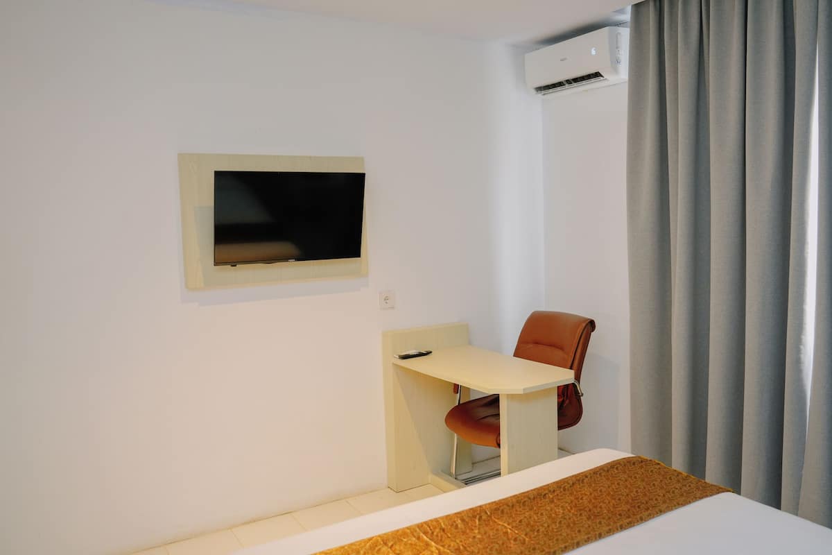 Quarto de hotel com cama, escrivaninha, cadeira, televisão e janela com cortinas.
