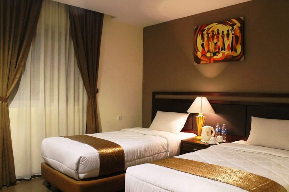Quarto de hotel com duas camas, um quadro na parede, uma luminária na mesinha de cabeceira e uma janela com cortinas.