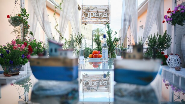 Uma mesa com tampo de vidro, rodeada por plantas em vasos e flores pendentes, com placas que dizem 'Opera' e 'Obelisk'.