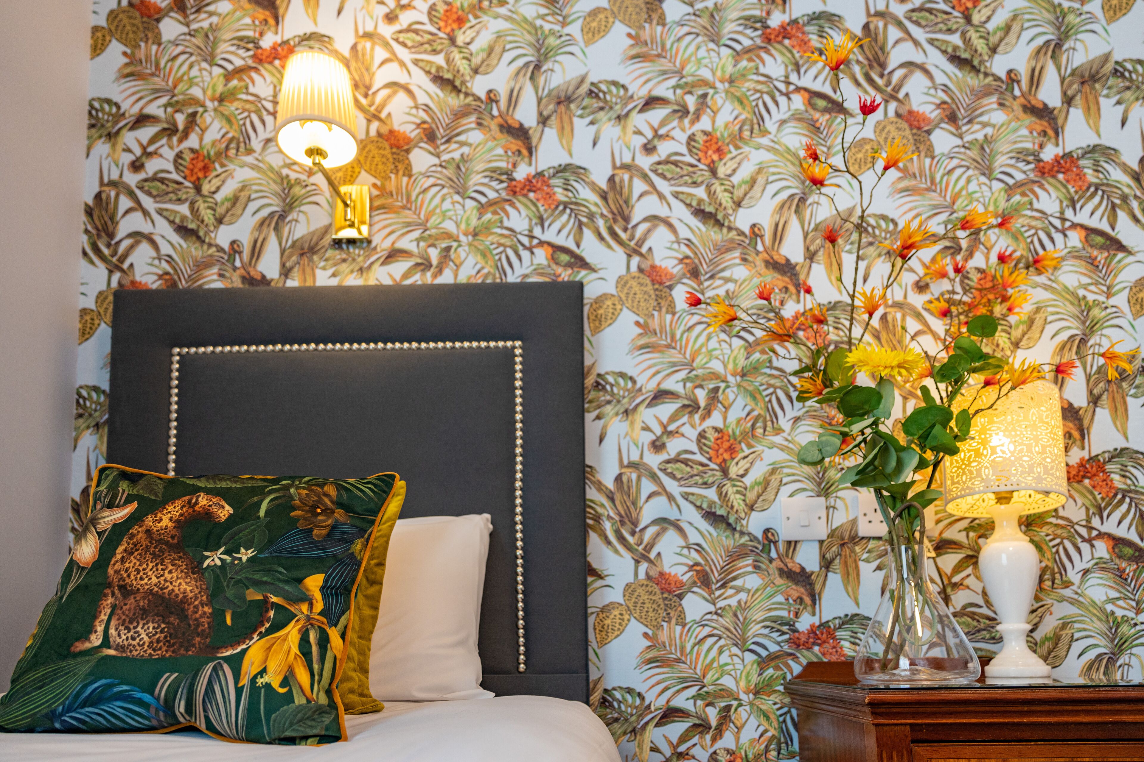 Quarto com papel de parede floral, almofada de estampa de leopardo, almofada branca, cabeceira escura e mesa de cabeceira com abajur.