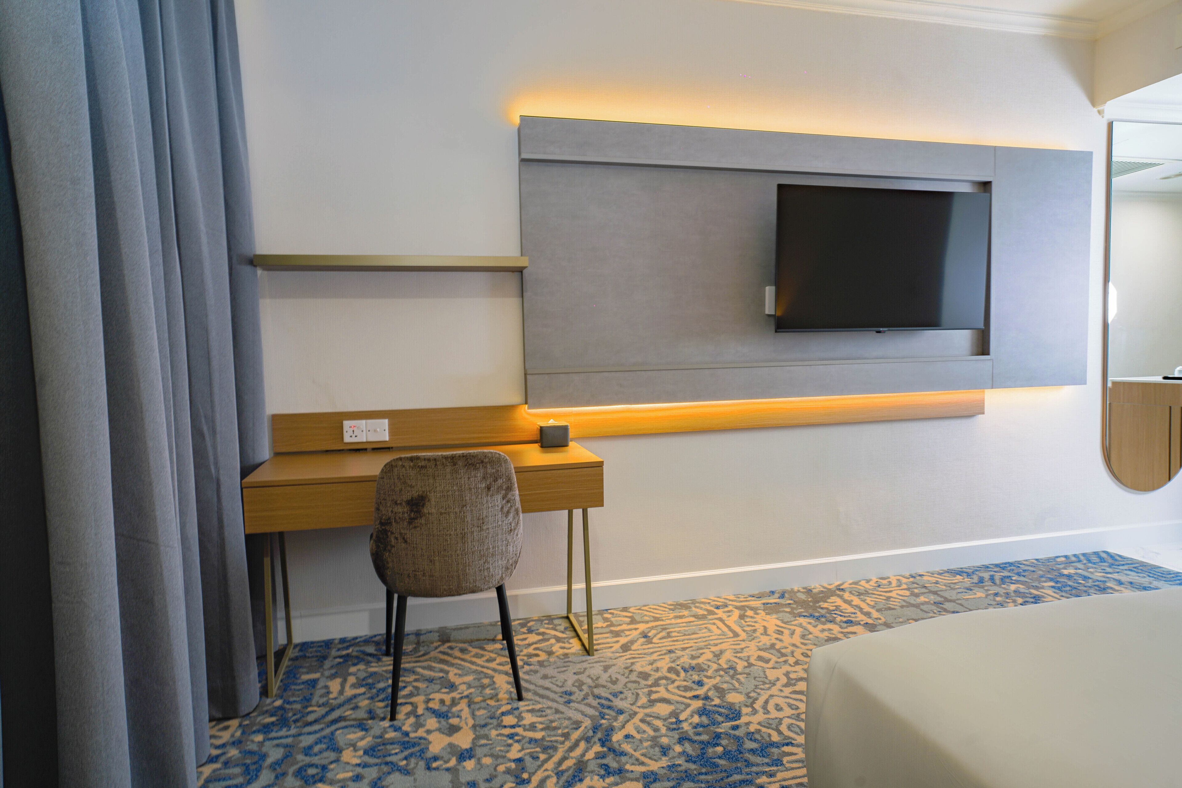 Quarto de hotel com cama, escrivaninha, cadeira, televisão fixada na parede e carpete estampado.