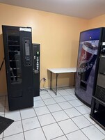 Quarto com máquina de vendas automática, uma mesinha e um frigorífico fixado na parede.