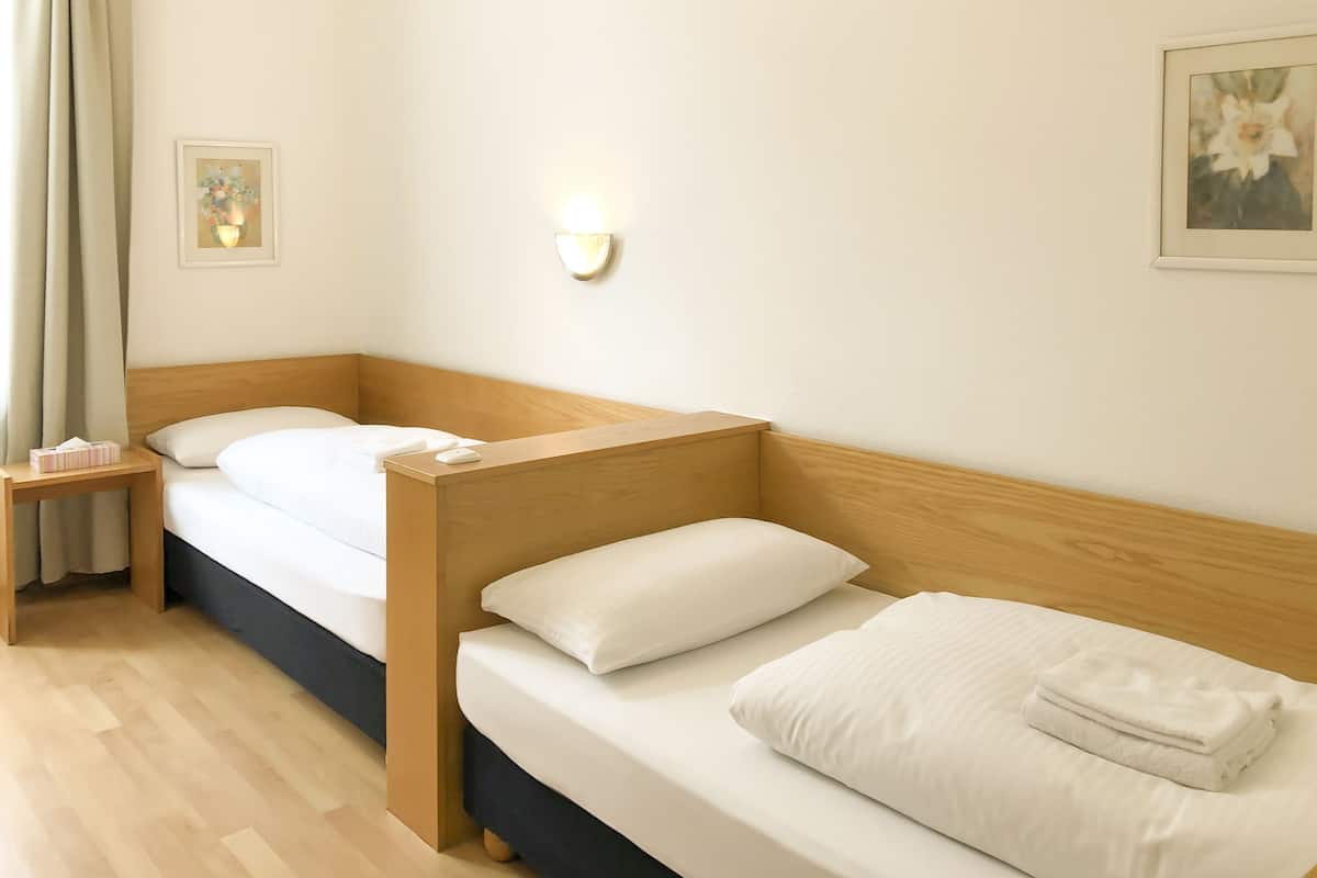 Quarto de hotel com estrutura de cama de madeira, roupa de cama branca, mesa de cabeceira com abajur e pequena mesa de apoio com caixa de lenços.