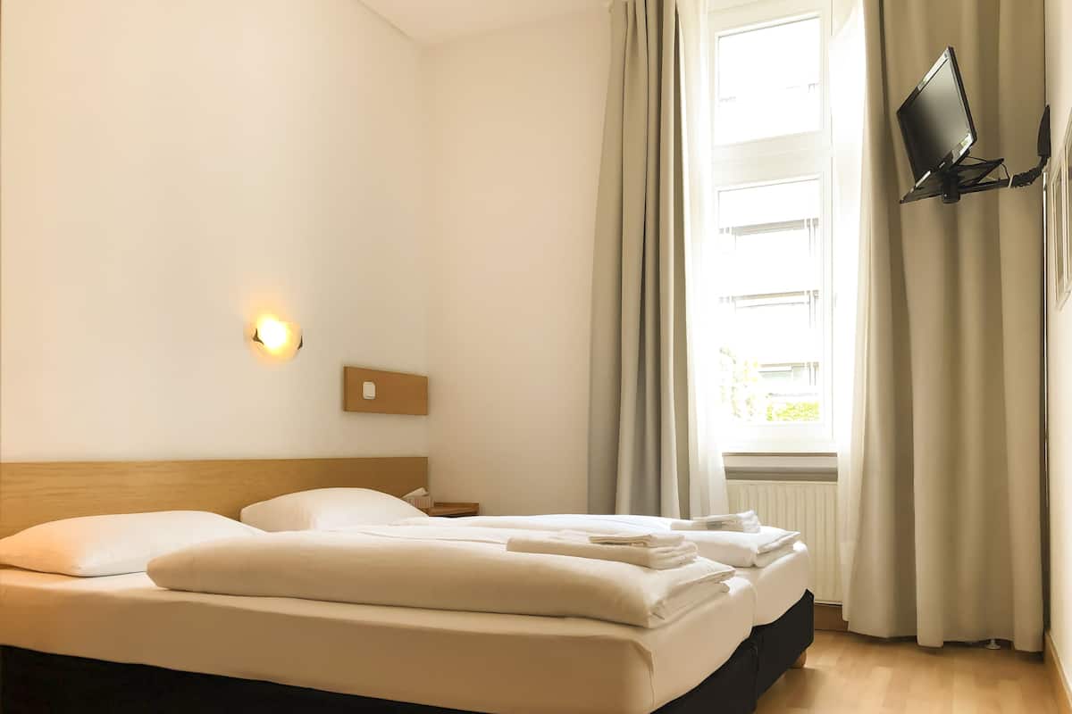 Quarto de hotel com cama, mesa de cabeceira, televisão na parede e janela com cortinas.
