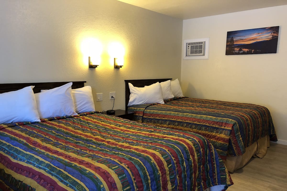Quarto de hotel com duas camas, cada uma com almofadas brancas e um edredom colorido listrado. Há um ar condicionado fixado na parede e um quadro emoldurado.