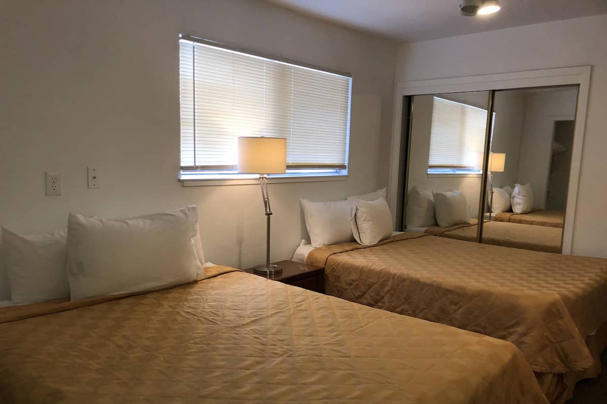 Quarto de hotel com duas camas, uma mesa de cabeceira com luminária, um espelho grande e uma janela com persianas.