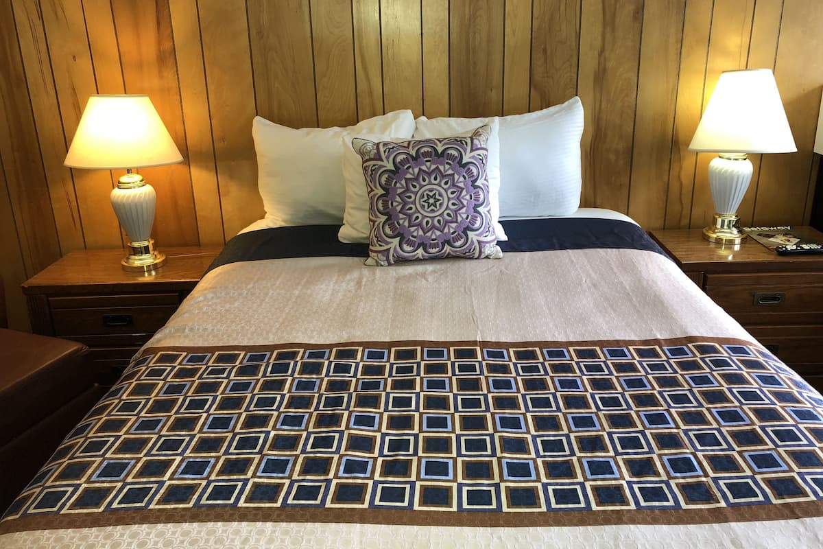 Quarto de hotel com parede revestida em madeira, uma cama grande com colcha estampada, duas luminárias de cabeceira e uma mesa de cabeceira com controle remoto.