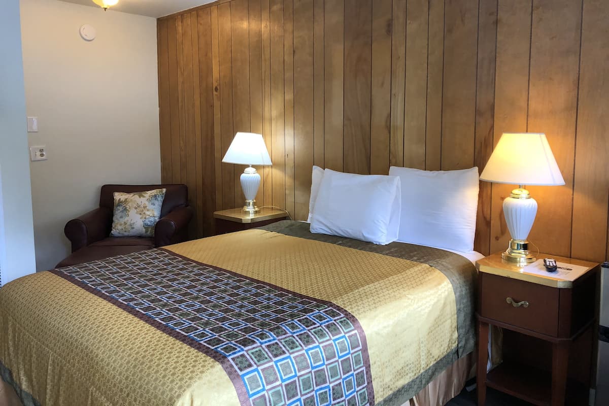 Quarto de hotel com cabeceira de madeira, cama com colcha estampada, duas mesas de cabeceira com luminárias, uma cadeira com almofada floral e um candeeiro fixado na parede.