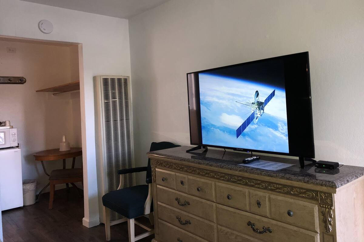 Quarto com televisão exibindo um satélite, uma cadeira de madeira, uma cômoda e um refrigerador branco.