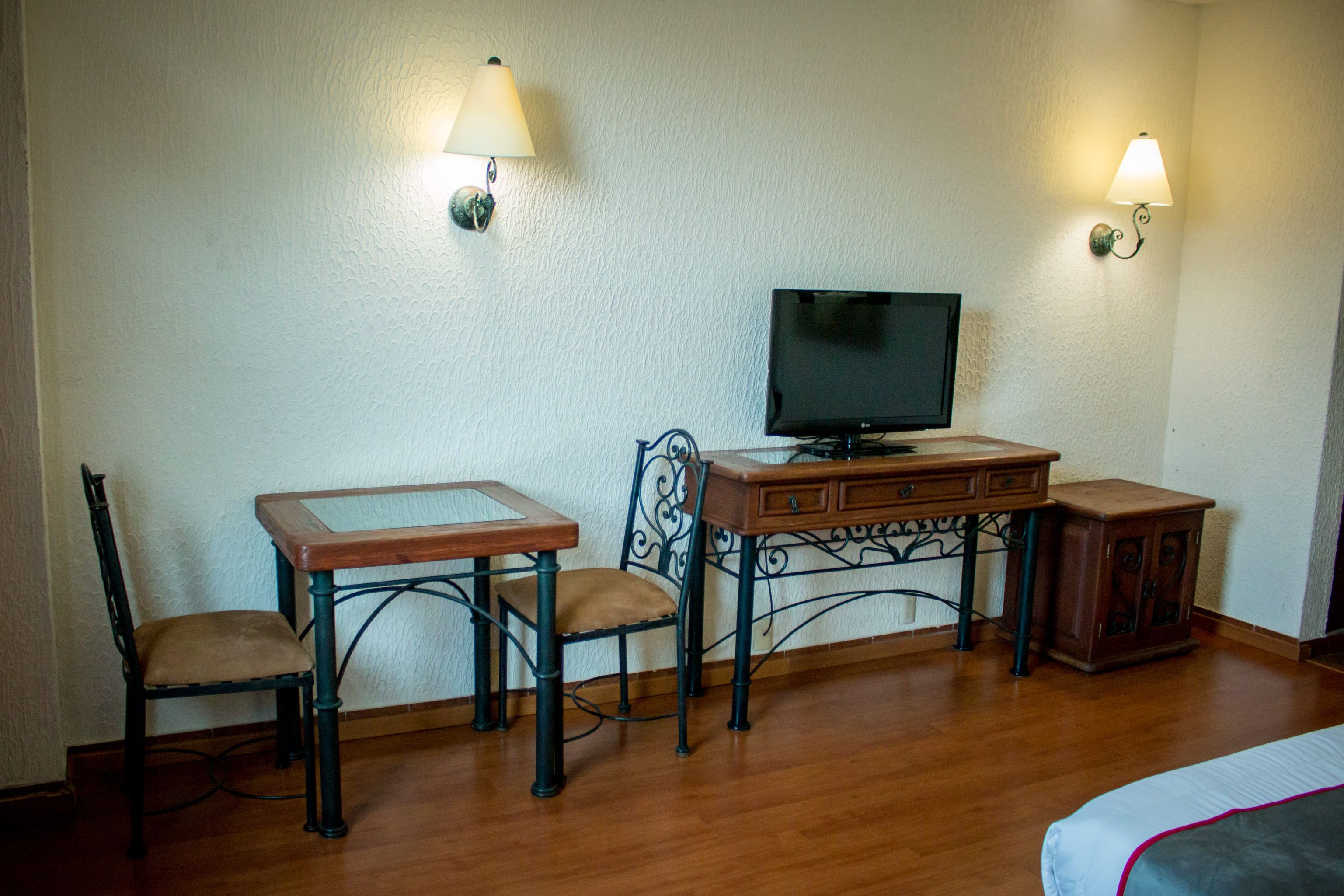 Um quarto com uma escrivaninha de madeira, uma cadeira, uma televisão e duas luminárias fixadas na parede.