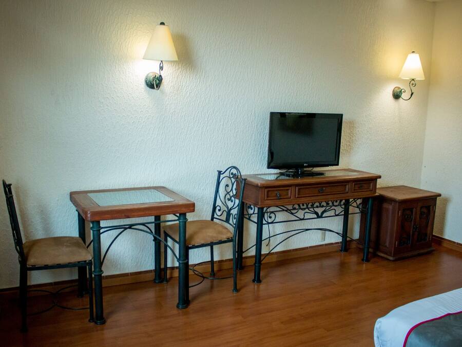 Um quarto com uma escrivaninha de madeira, uma cadeira, uma televisão e duas luminárias fixadas na parede.