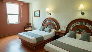 Quarto de hotel com duas camas, uma cadeira, uma mesa de cabeceira e uma luminária fixada na parede.