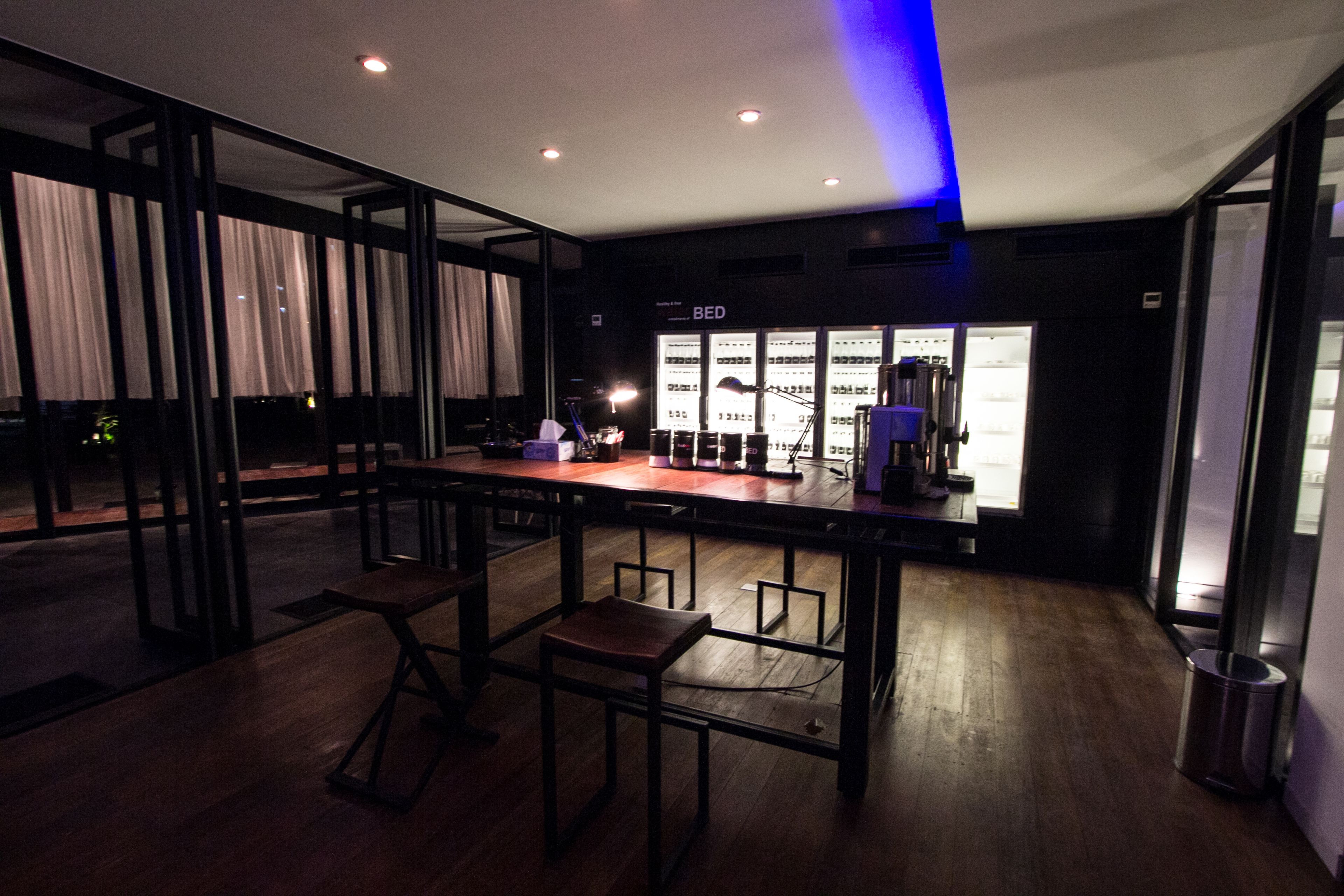 Uma área de bar moderna com um balcão comprido, vários bancos e uma geladeira com a etiqueta 'BED' ao fundo.