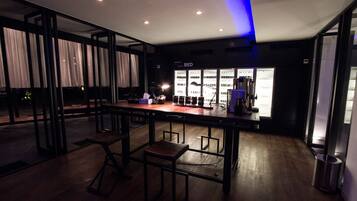 Uma área de bar moderna com um balcão comprido, vários bancos e uma geladeira com a etiqueta 'BED' ao fundo.