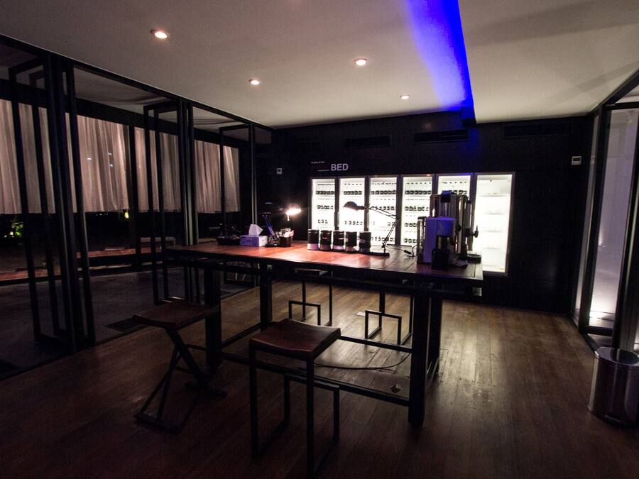 Uma área de bar moderna com um balcão comprido, vários bancos e uma geladeira com a etiqueta 'BED' ao fundo.