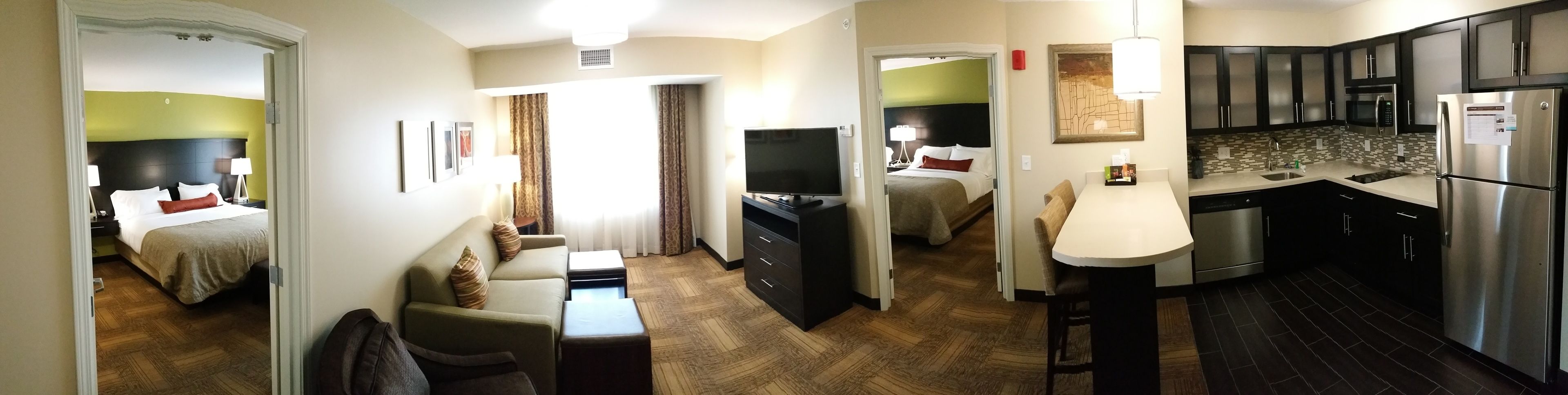 Quarto de hotel com cama, sofá, televisão, escrivaninha e cozinha compacta.