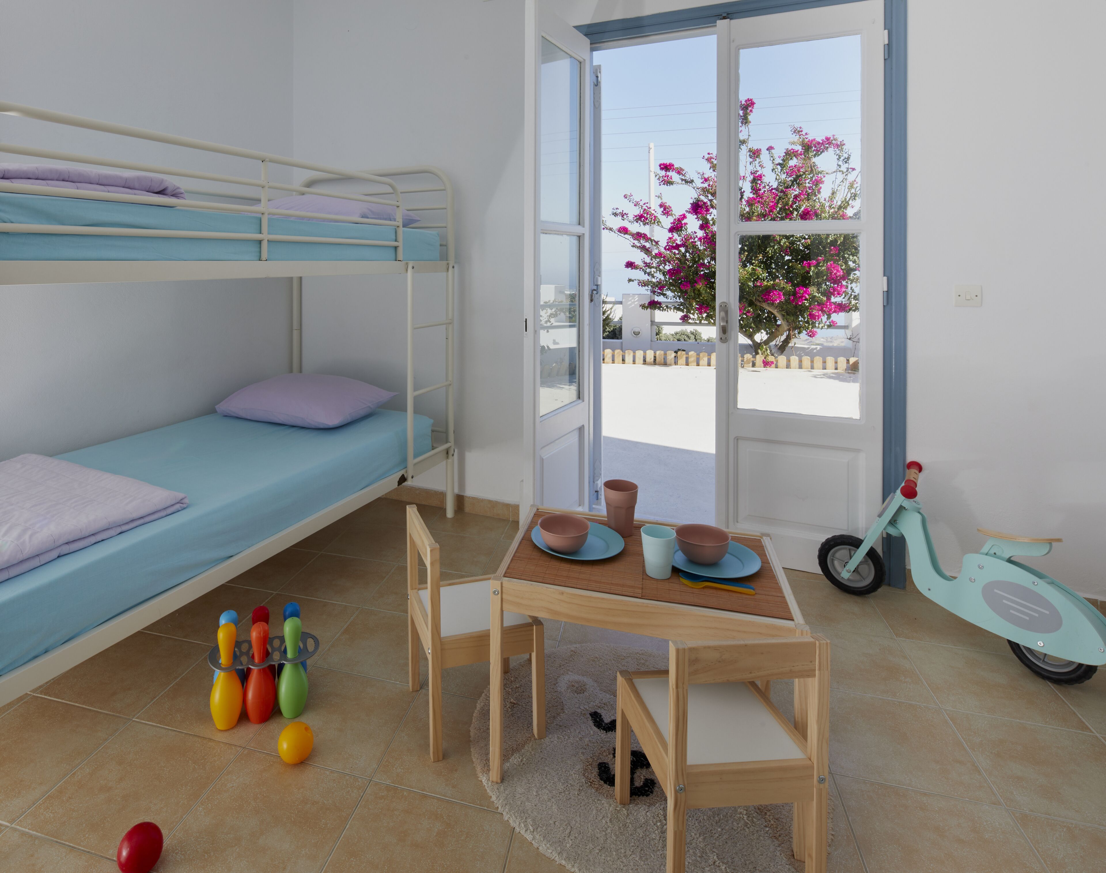 Quarto com beliches, uma mesa com tigelas, uma scooter e uma janela florida.