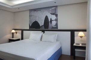 Quarto de hotel com uma cama grande, duas mesas de cabeceira com luminárias, uma cabeceira e uma obra de arte na parede.