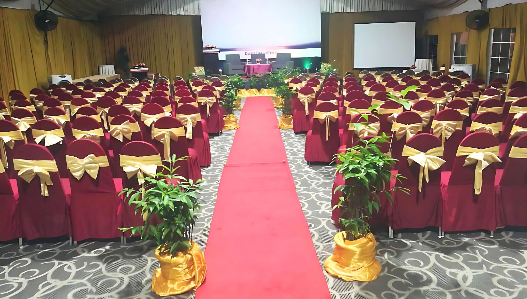 Uma sala de conferências com fileiras de cadeiras, tapete vermelho e plantas em vasos.