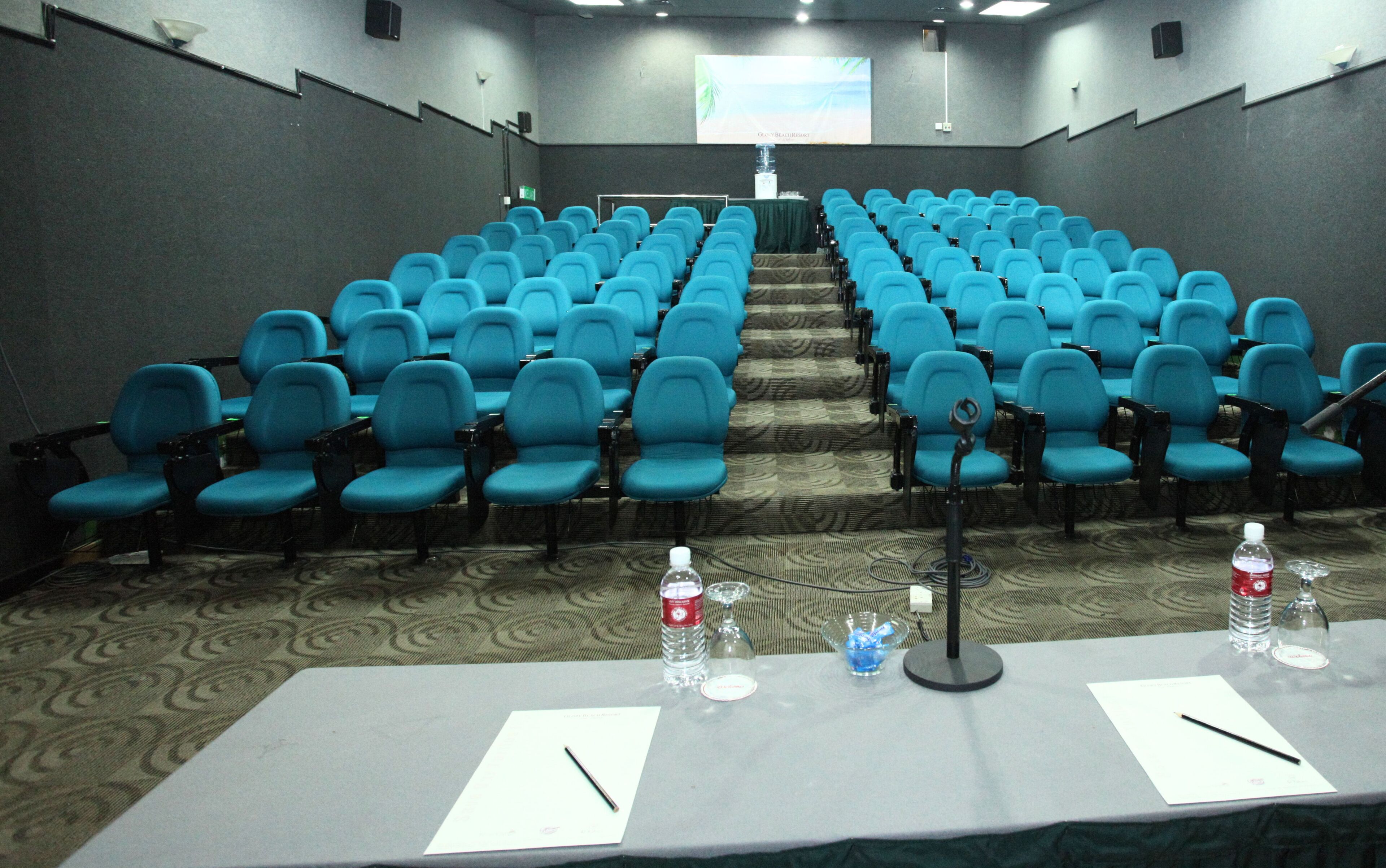 Uma sala de conferências com fileiras de cadeiras azuis, piso coberto de carpete, uma tela de projetor na frente e mesas com garrafas de água e canetas.