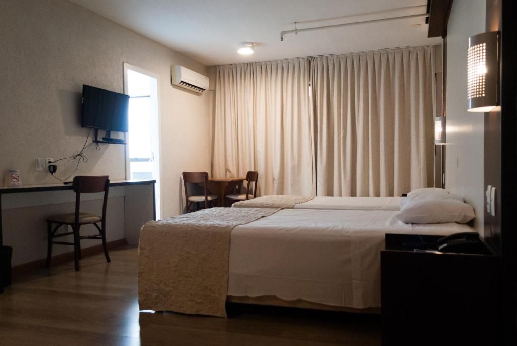 Quarto de hotel com duas camas, uma escrivaninha com cadeira, televisão fixada na parede e janela com cortinas.