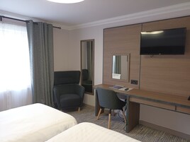 Quarto de hotel com duas camas, uma escrivaninha com cadeira, televisão de tela plana, espelho e janela com cortinas.