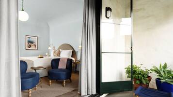 Quarto de hotel com uma cama, duas poltronas azuis, uma mesa pequena, uma planta em vaso e vista para o exterior.