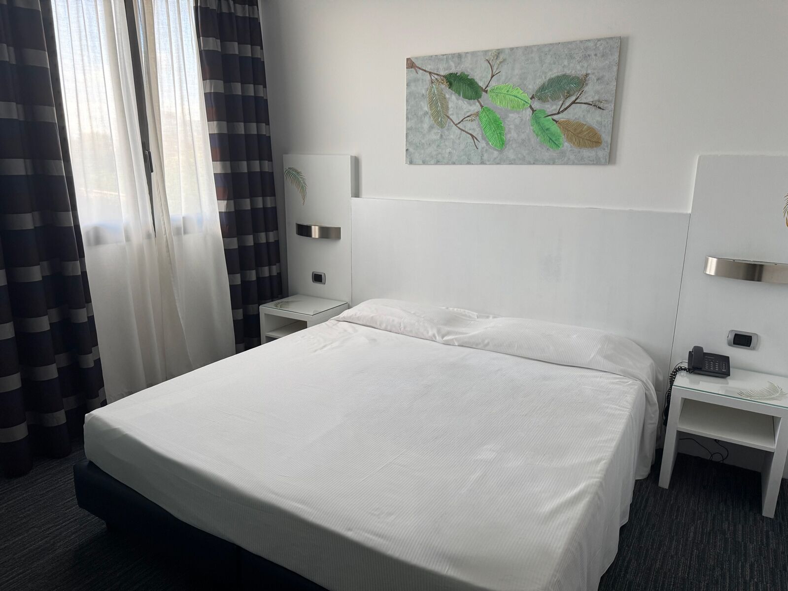 Quarto de hotel com uma cama grande, uma mesinha de cabeceira com telefone e uma obra de arte na parede.