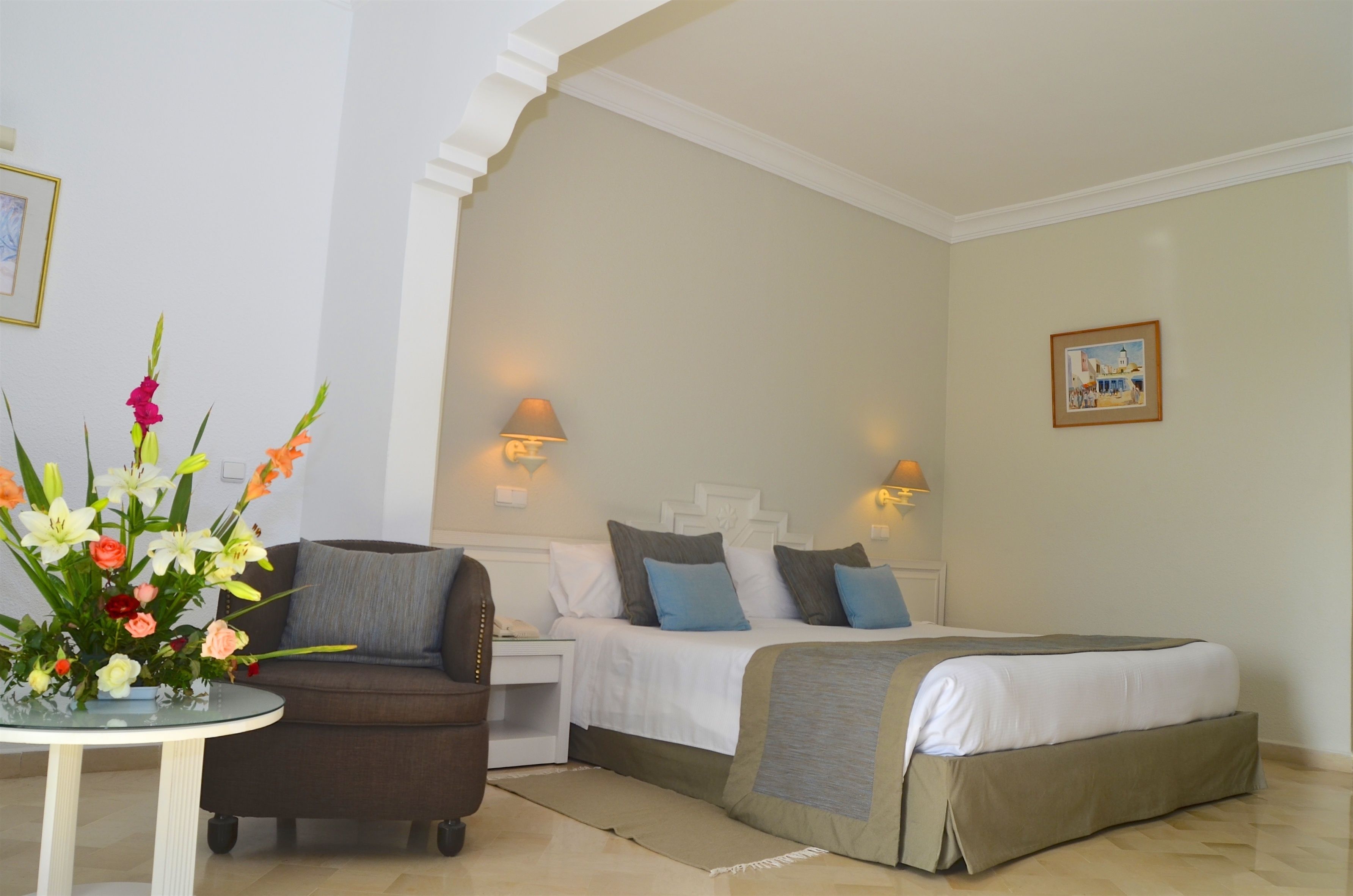 Quarto de hotel com uma cama, uma cadeira, uma mesinha com flores e um quadro na parede.