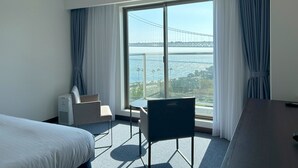 Quarto de hotel com uma grande janela que oferece vista para uma ponte e o mar. Há uma cama, uma escrivaninha e uma cadeira.