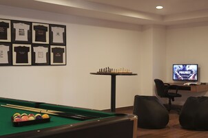 Uma mesa de bilhar com bolas, taco de bilhar, televisão exibindo uma partida e uma parede com camisetas emolduradas.