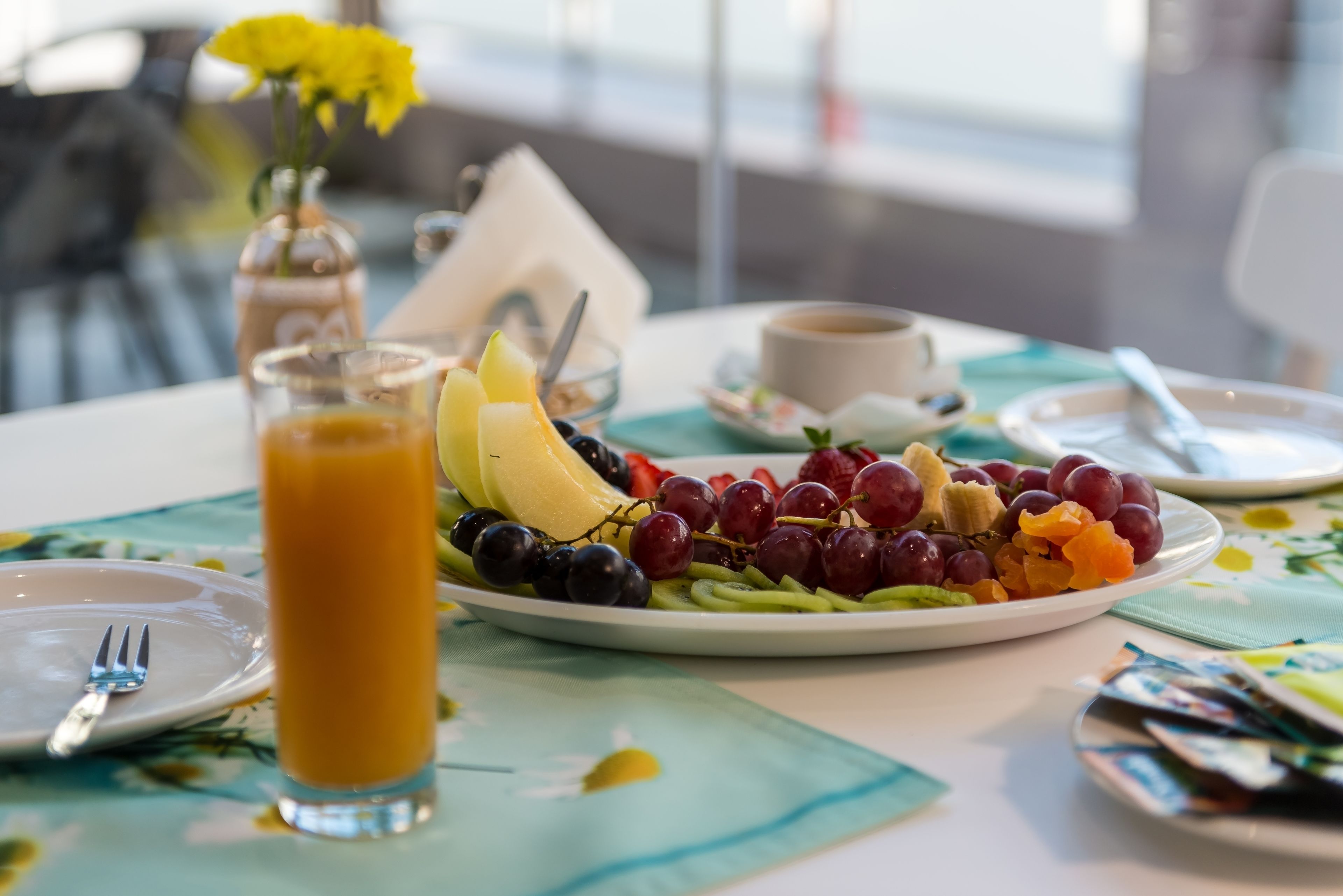 Disposição para o pequeno-almoço com uma tábua de frutas, um copo de sumo de laranja e uma chávena de café sobre uma mesa com toalha floral.